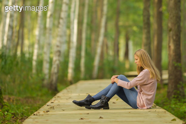 Sad young adult woman sitting alone on wooden trail at birch tree ...