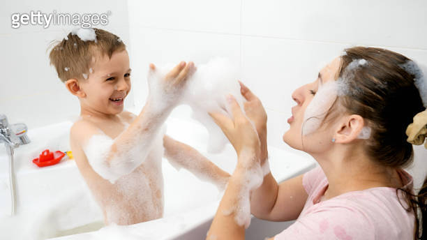 Funny mother with son splashing water in bath and throwing soap suds ...