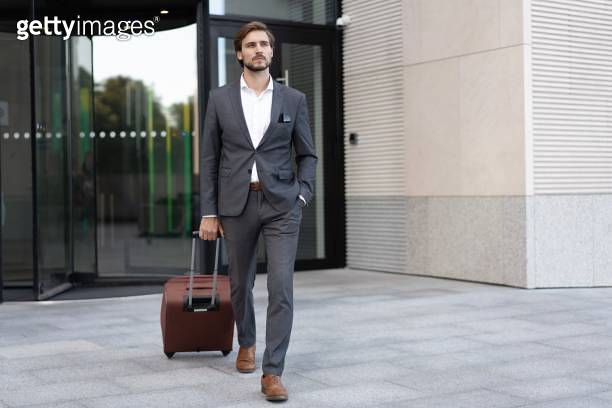 Smiling and good looking manager with suitcase walking outside modern ...