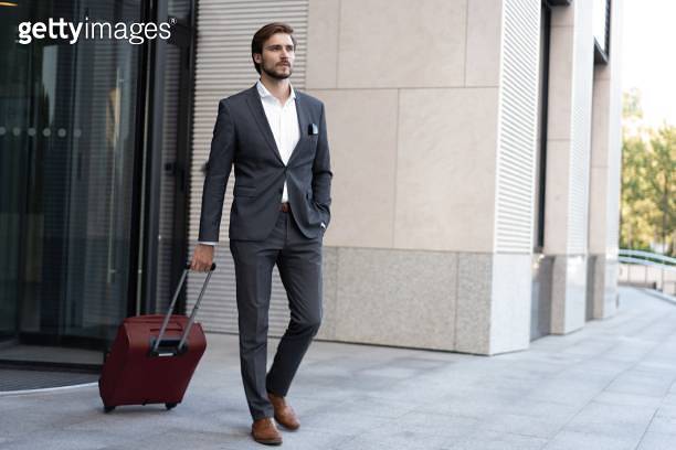 Smiling and good looking manager with suitcase walking outside modern ...