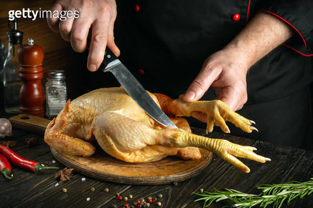 The chef prepares to cut raw rooster with a knife on a kitchen cutting ...