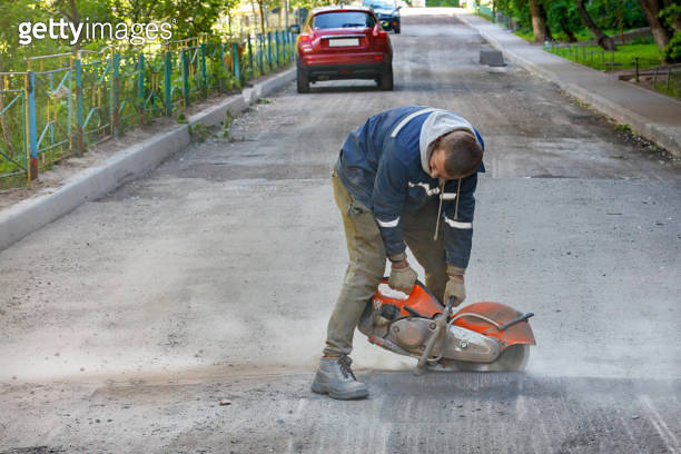 A worker cuts worn-out asphalt with a petrol cutter in a cloud of dust ...