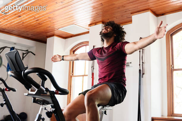 Young middle eastern man with arms outstretched on exercise bike at the ...