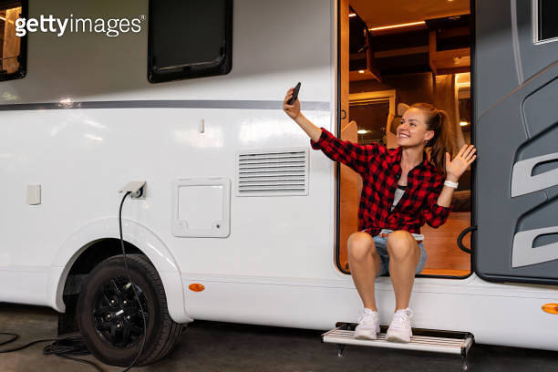 Camper van travel lifestyle, woman takes a selfie using her cellphone while her electric camper ...