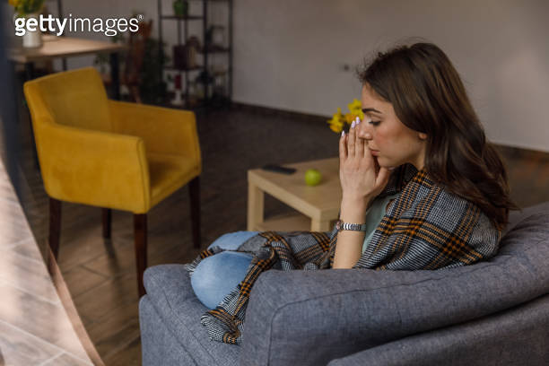 Young woman sitting on the sofa, wrapped in a blanket, fidgeting her ...