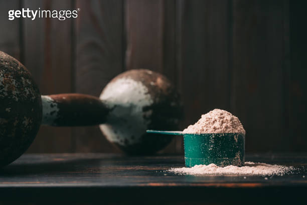 Chocolate whey protein powder in measuring spoon, old rusty dumbbell on ...