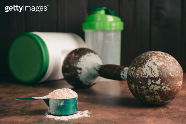 Chocolate whey protein powder in measuring spoon, old rusty dumbbell ...