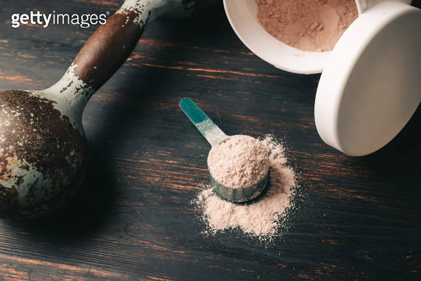 Chocolate whey protein powder in measuring spoon, old rusty dumbbell ...