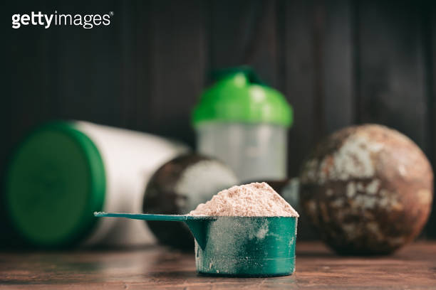 Chocolate whey protein powder in measuring spoon, old rusty dumbbell ...
