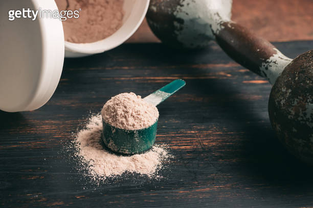 Chocolate whey protein powder in measuring spoon, old rusty dumbbell ...