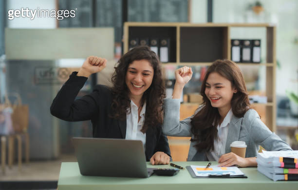 Two businesswoman showing joy, raising hands at workplace, smiling and ...