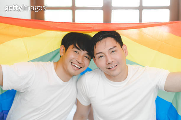 Happy Asian LGBT couple, sitting on the sofa holding and waving rainbow ...