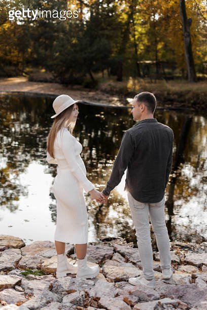 Rear view of stylish young couple in love holding hands by the pond at ...