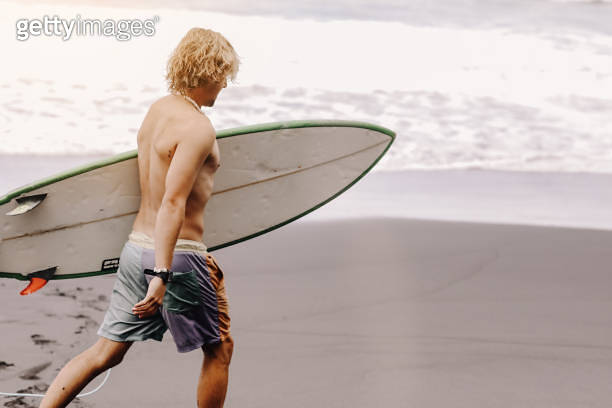 Fit young surfer man with curly blond hair with surfboard goes by the ...