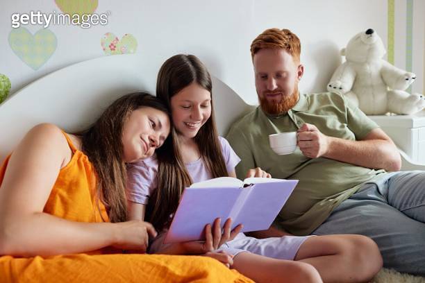 Father and two daughters are reading a book together on a bed, with the ...