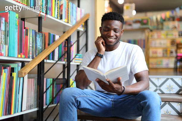 In the university library, a young man sits among bookshelves, smiling ...
