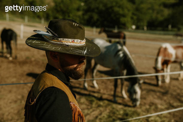 Side view of rancher with hat at ranch with horses grazing grass. 이미지 ...