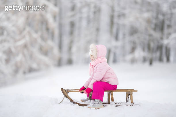 Happy little girl in pink warm clothes sitting on sledge and sledding ...