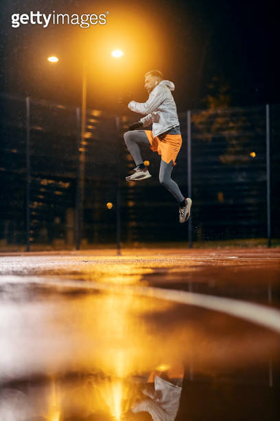 A fit urban sportsman is jumping high on sports ground on rainy cold ...