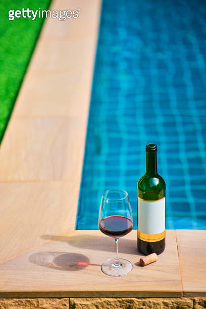 A bottle of red wine and a glass of wine are set on the edge of a clear ...