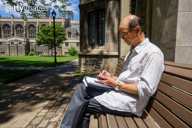 A male professor sitting outside on the university campus, reading ...