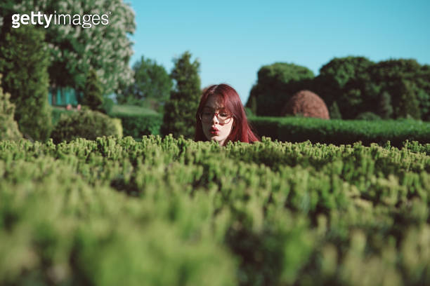 Teenage girl with glasses peeking over green bushes in a park. Outdoor ...