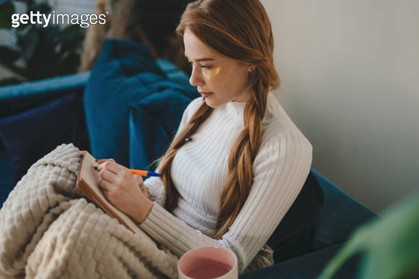 Young redheaded woman sitting on the sofa wraped in a blanket writing ...