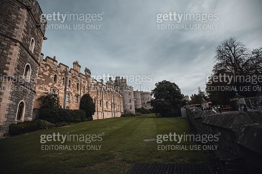 A stone castle wall with small windows overlooks a grassy field and a ...