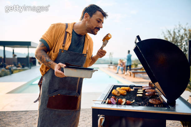 Young man grilling food on a barbecue during pool party with friends ...