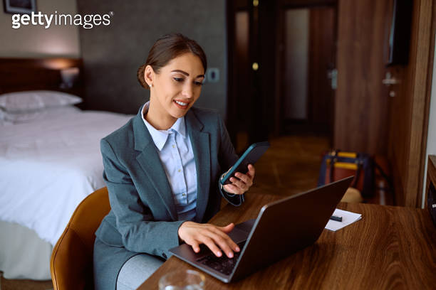 Smiling businesswoman using cell phone while working on laptop in hotel ...