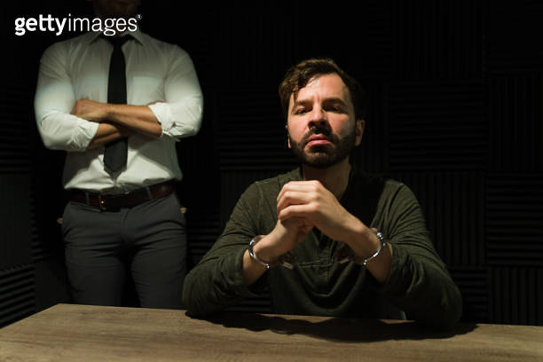 A focused detective stands behind as a suspect sits tensely under a ...