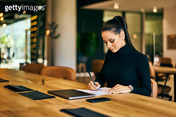 A focused smiling woman making some plans, writing on paper, at the co ...