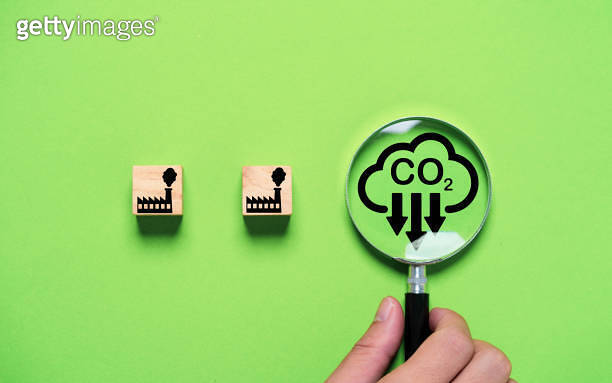 Hand holding magnifier glass with CO2 reducing icon and black factory for decrease CO2 emission ...