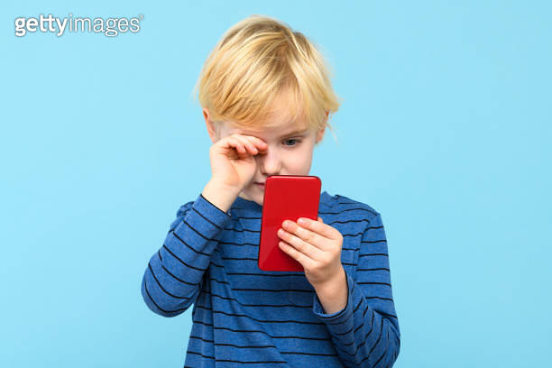 Young child holding smartphone, watching the screen, rubbing his eyes ...