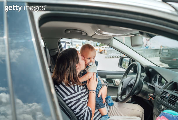 Mother and baby sit in parked car with baby playfully holding steering ...