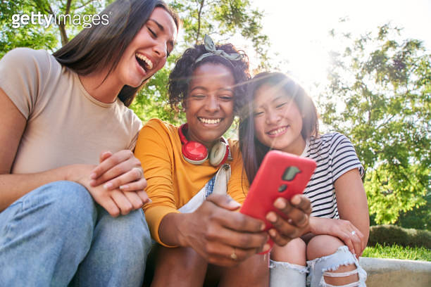 Group joyful friends using mobile sitting outdoor. Three laughing ...