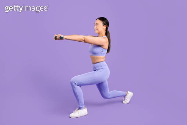 Asian woman in lunge holding dumbbell with extended arms on purple ...